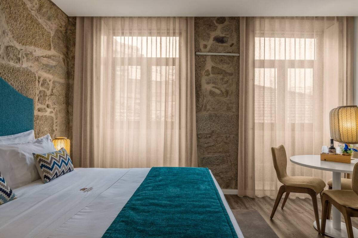Quarto de hotel com parede em pedra decorativa, cama com manta azul-turquesa, mesa de jantar redonda com cadeiras e uma luminária.