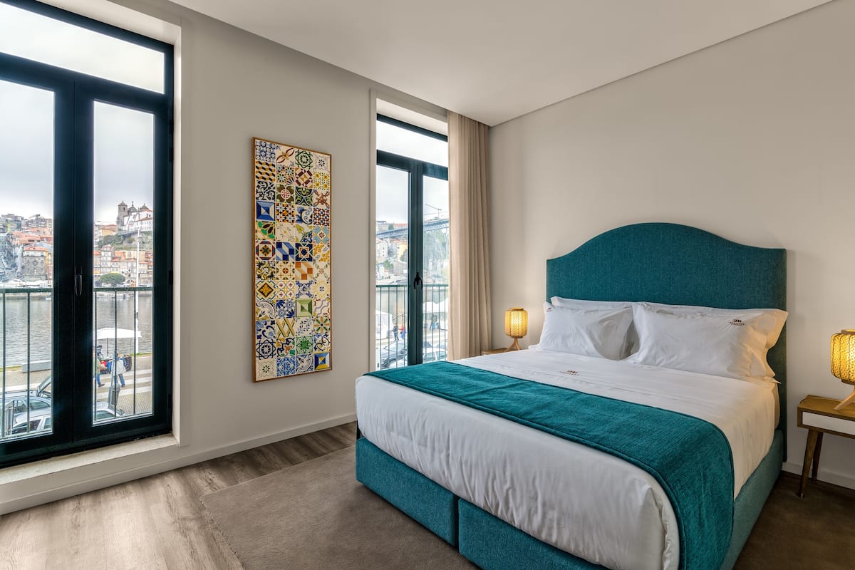 Quarto de hotel com uma cama grande, vista da cidade por grandes janelas e uma obra de arte colorida na parede.