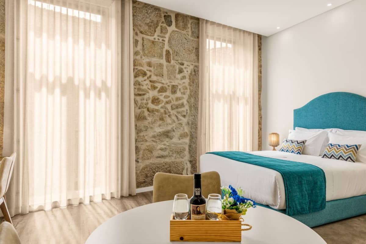 Quarto com parede de pedra, cama com cabeceira azul-turquesa e mesa de jantar com uma garrafa e copos.