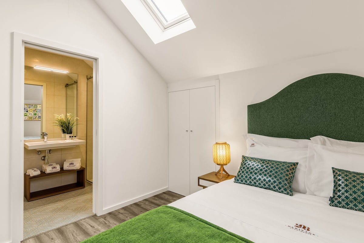 Quarto com claraboia, cama com almofadas verdes, abajur e vista para o banheiro.