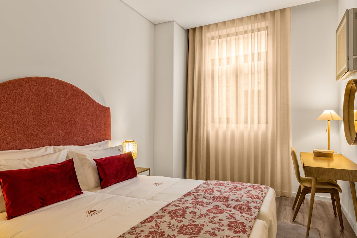 Quarto de hotel com uma cama, escrivaninha, cadeira, abajur e televisão.