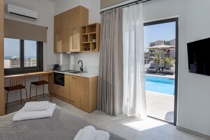 Quarto de hotel moderno com cozinha compacta, cama com roupa de cama branca, uma janela grande e vista para uma piscina e edifícios.