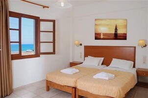 Uma cama de casal com duas almofadas, uma mesa de cabeceira, um quadro, uma janela com cortinas e vista para o mar.