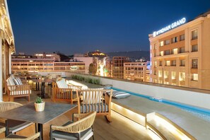 Terraço no telhado com área de estar externa, piscina e vista para a cidade à noite.