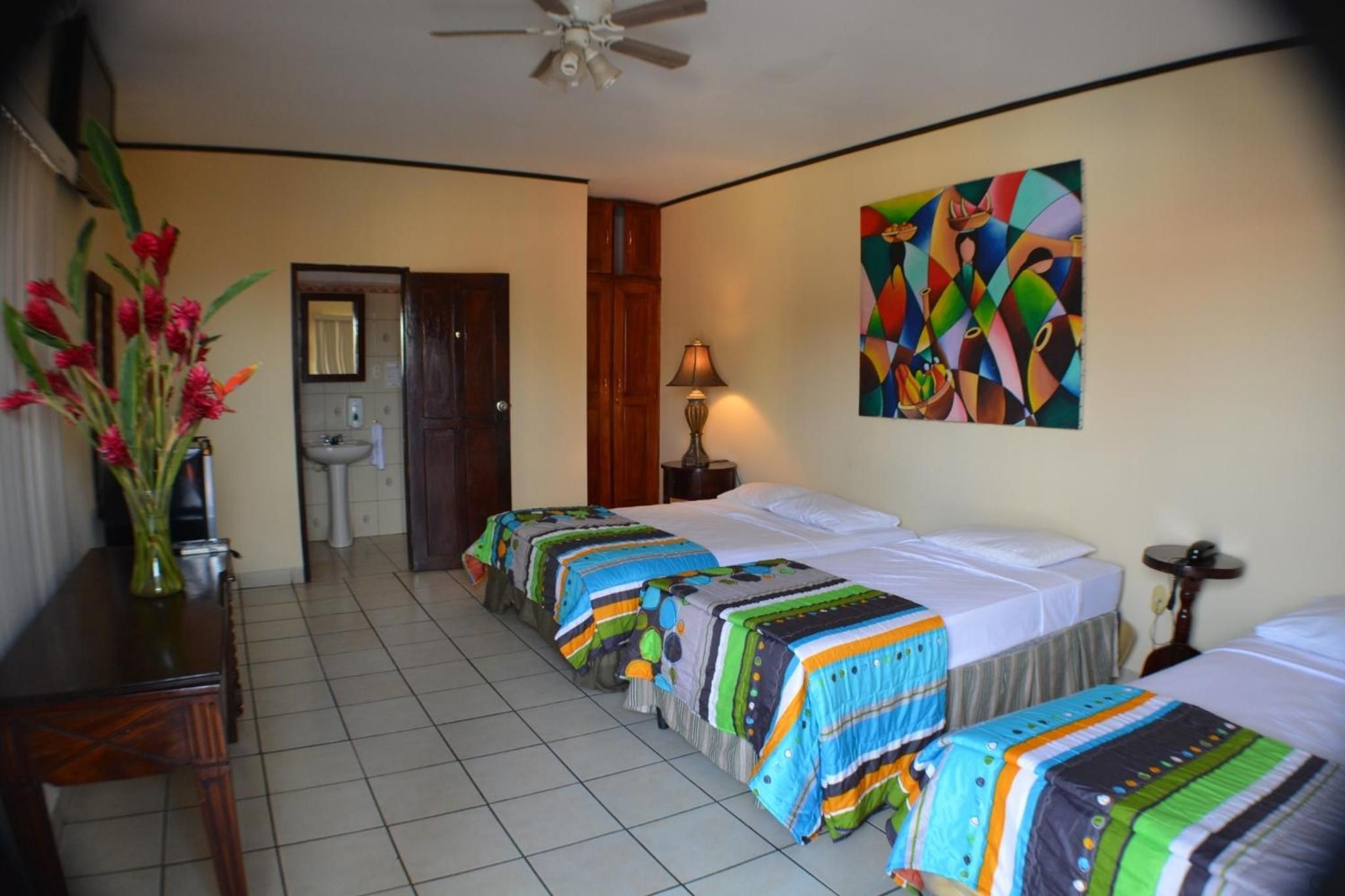 Quarto de hotel com duas camas, uma escrivaninha, uma cadeira, um vaso com flores, um quadro na parede e um ventilador de teto.