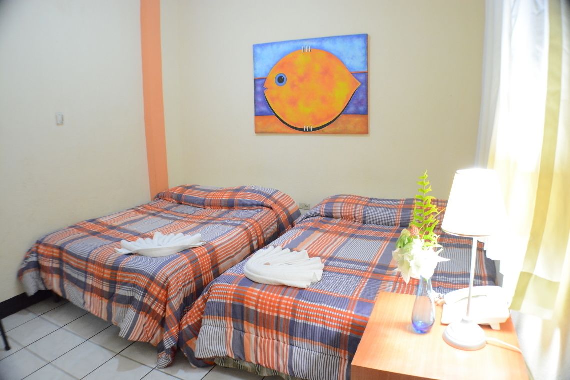 Quarto de hotel com duas camas, um quadro na parede, uma luminária e uma planta no criado-mudo.