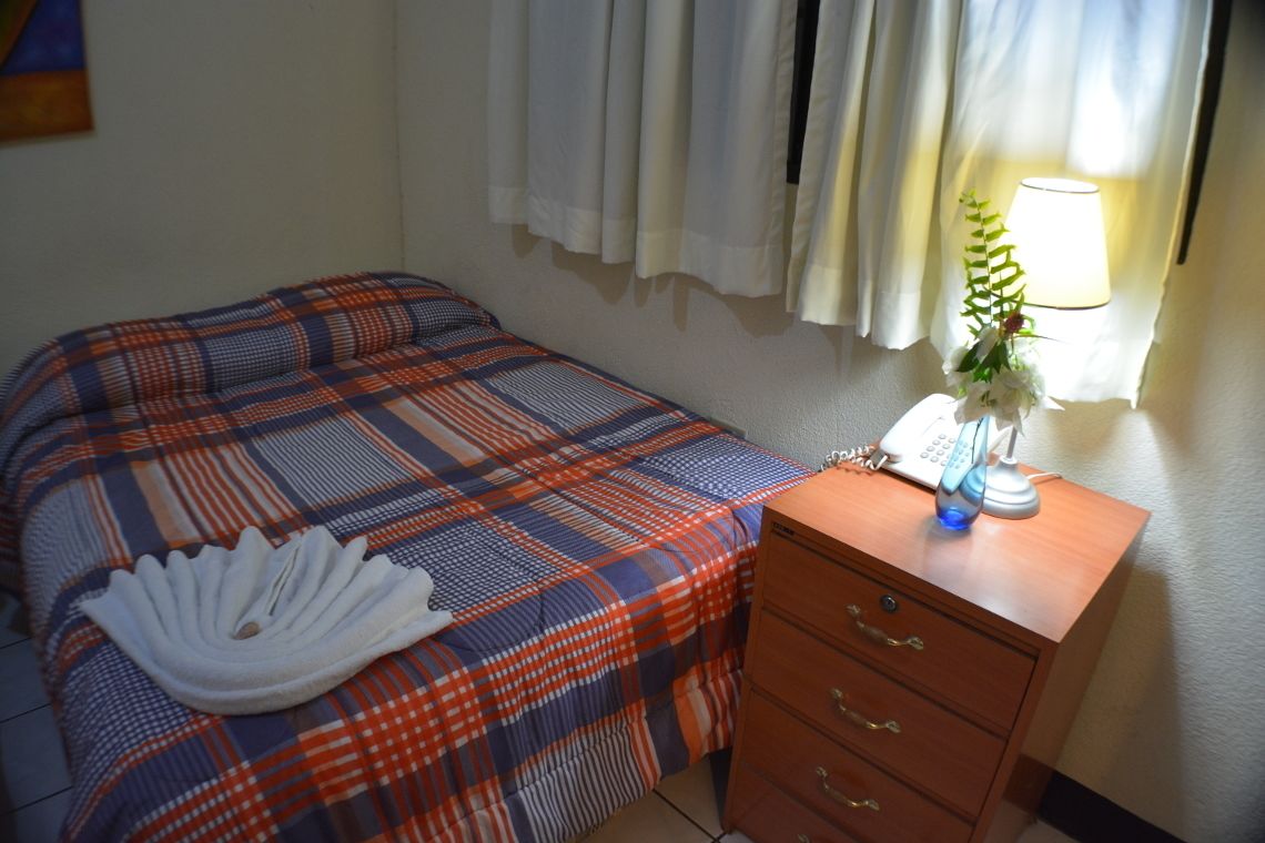 Quarto de hotel com cama, mesa de cabeceira com abajur, telefone e um objeto decorativo.