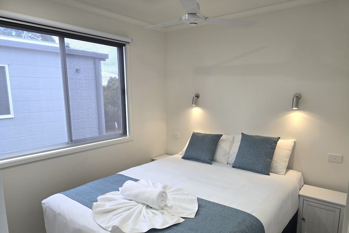 Cama bem arrumada com toalha, janela com persianas e ventilador de teto.