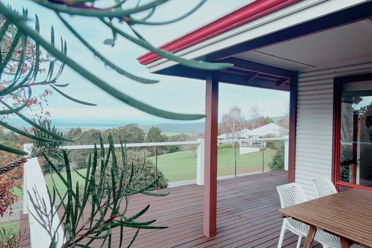 Uma área externa coberta com um deck de madeira, cadeiras brancas e vista para uma paisagem verde.