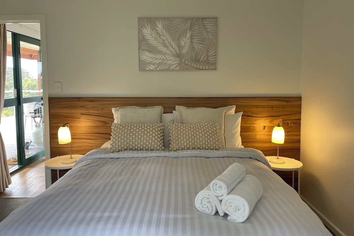 Quarto de hotel com uma cama, duas luminárias de cabeceira, um painel de cama de madeira e uma obra de arte com padrão de folhas.