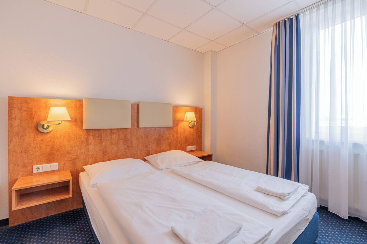 Quarto de hotel com uma cama de casal, duas mesas de cabeceira, uma luminária na parede, um espelho e uma janela com cortinas.