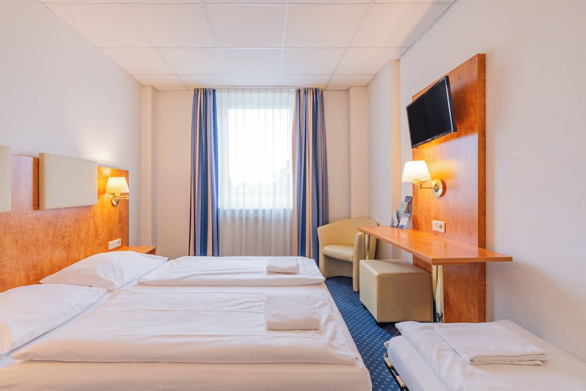 Quarto de hotel com duas camas, uma escrivaninha com televisão, uma cadeira e uma janela com cortinas.