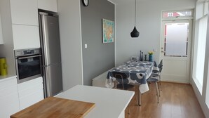 Uma cozinha moderna com forno embutido e frigorífico, uma mesa de jantar com toalha de mesa estampada e uma parede cinzenta com um mapa.