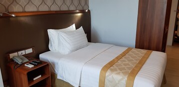 Quarto de hotel com uma cama, uma mesa de cabeceira de madeira com telefone e controles remotos, e um painel de cabeceira estampado.