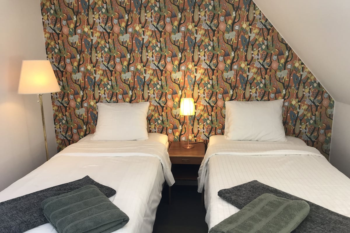 Quarto com duas camas, papel de parede estampado, mesa de cabeceira com abajur e toalha verde em cada cama.