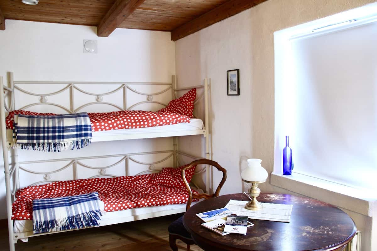 Quarto com duas camas de beliche, uma cadeira de madeira, uma mesa redonda com abajur e uma janela com vista.