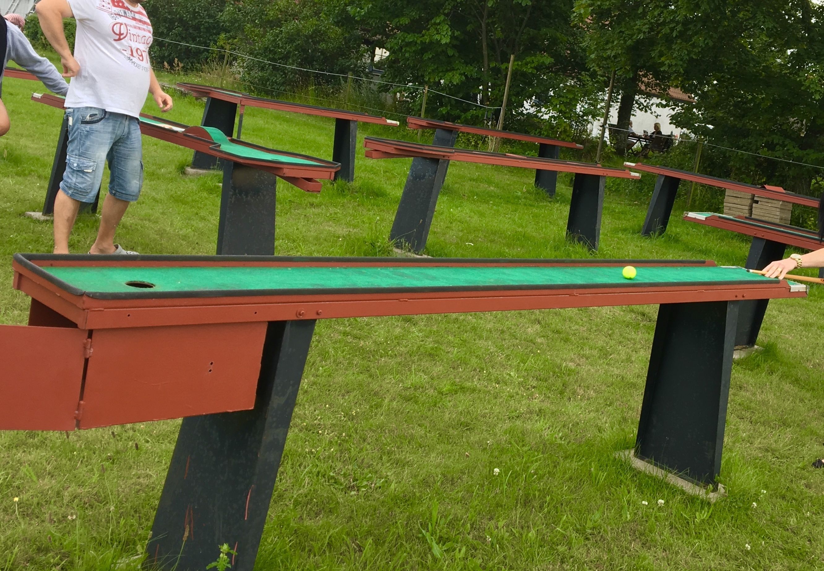 Uma mesa de bilhar com superfície em feltro verde e borda vermelha, cercada por grama e árvores.
