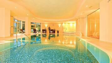 Piscina interna com chão em mosaico, área de estar com cadeiras e parede de vidro de um lado.