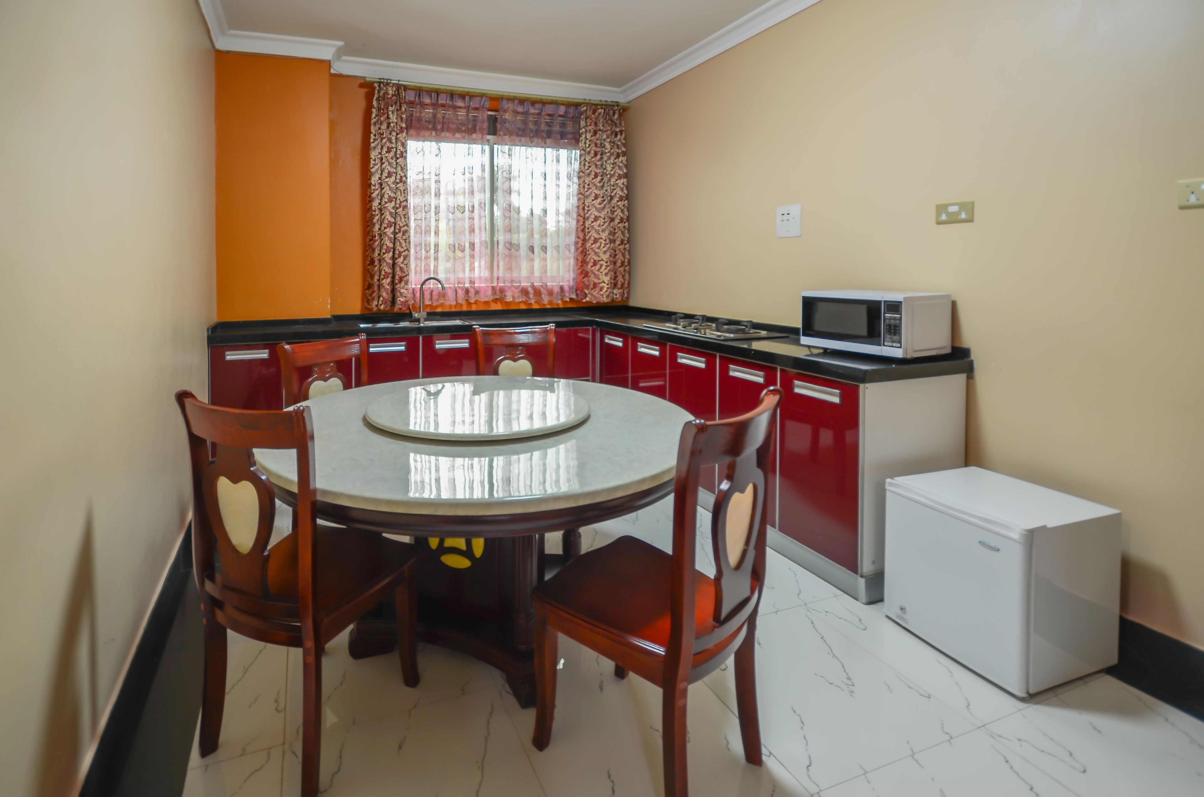 Uma cozinha compacta com uma mesa de jantar redonda, armários vermelhos, micro-ondas e uma geladeira pequena.