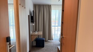 Quarto de hotel com piso de madeira, uma escrivaninha com cadeira, um pequeno banquinho e uma TV fixada na parede, além de uma janela com cortinas.