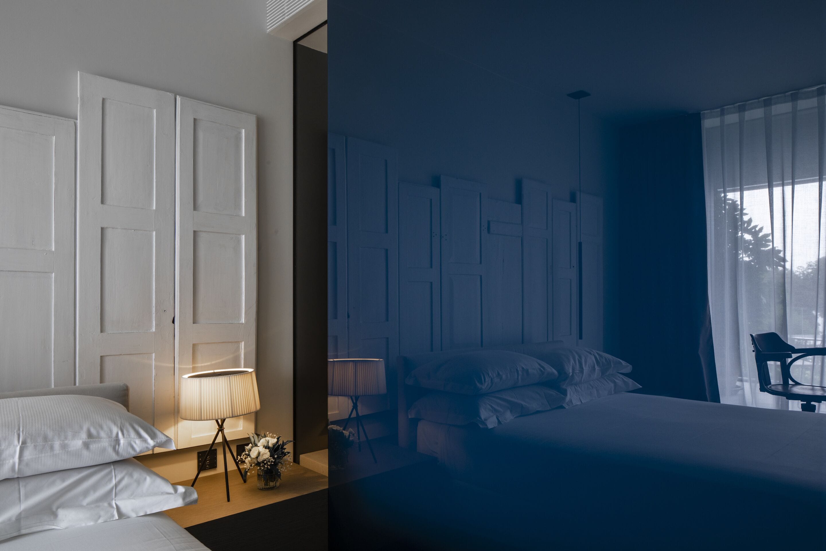 Quarto de hotel com cama, mesa de cabeceira com abajur e cadeira perto da janela.