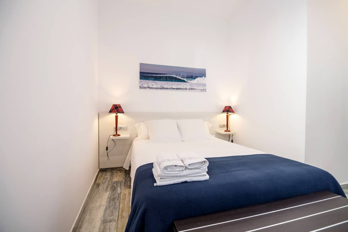 Quarto de hotel com cama, almofadas brancas, colcha azul, toalha dobrada, pintura de uma onda e duas luminárias de cabeceira.