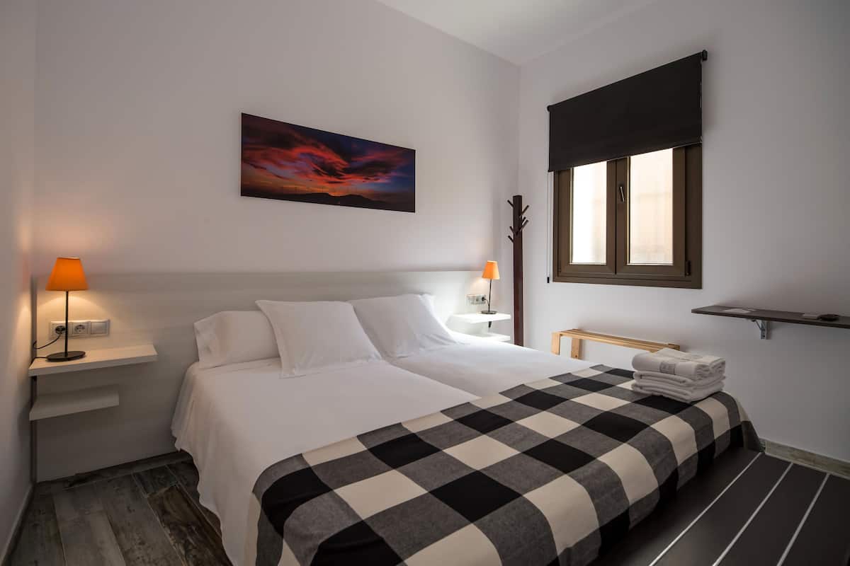 Quarto de hotel com uma cama, duas luminárias de cabeceira, um quadro, uma janela com persiana e uma pequena prateleira.
