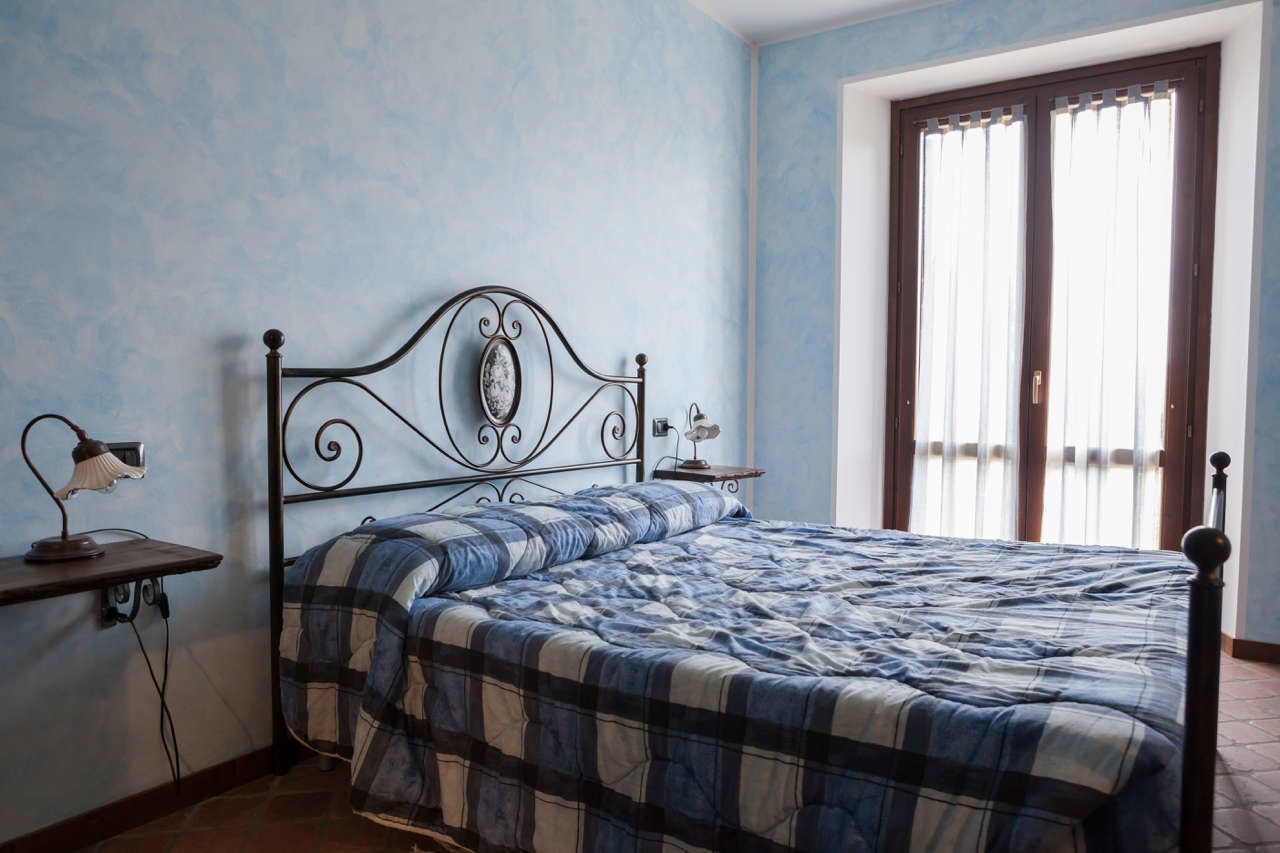 Quarto com estrutura de cama de metal, colcha xadrez azul e branca, mesa de cabeceira com abajur e janela com cortinas.