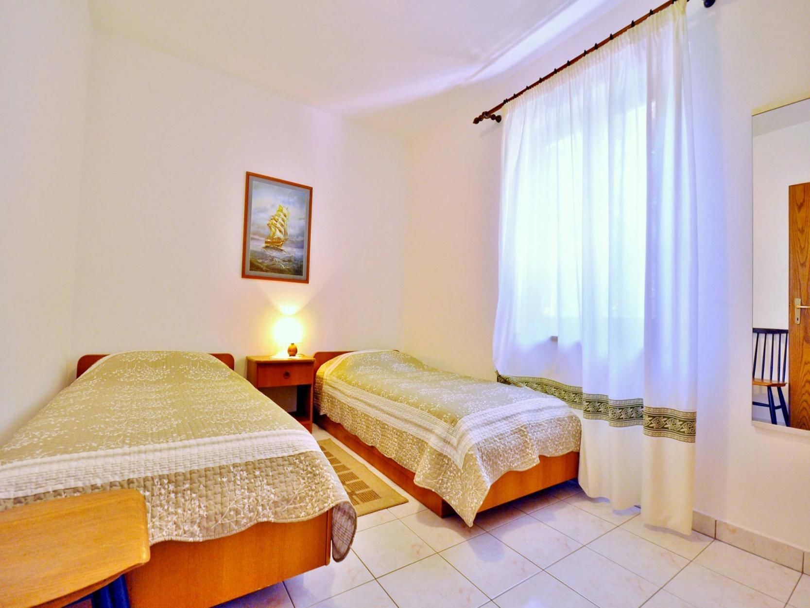 Quarto com duas camas de solteiro, uma mesa de cabeceira com abajur, um quadro na parede e uma janela com cortinas leves.