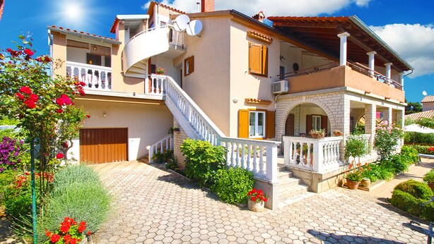Uma casa de dois andares com varanda, escadaria e um jardim com flores vermelhas.