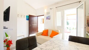 Uma sala de estar com sofá, duas poltronas, mesa de centro de mármore, televisão na parede e porta que dá para área externa.