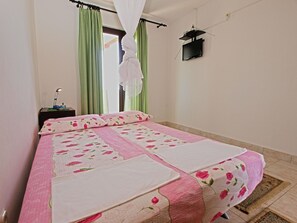 Quarto com cama, cortinas verdes, dossel branco, mesa de cabeceira com abajur e TV fixada na parede.