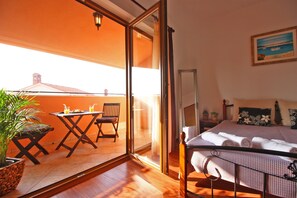 Quarto com cama, mesa de cabeceira, planta em vaso e vista para a varanda externa.