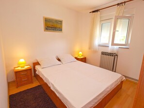 Quarto com cama, duas mesas de cabeceira com luminárias, radiador, janela com cortinas e um quadro na parede.