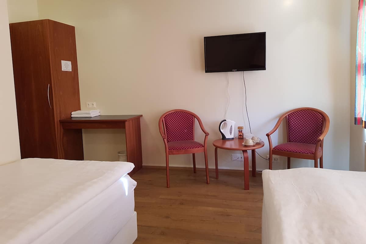 Quarto de hotel com duas camas de solteiro, um guarda-roupa de madeira, uma pequena escrivaninha, uma televisão fixada na parede e duas cadeiras vermelhas com estampa ao redor de uma mesinha com chaleira e xícaras.