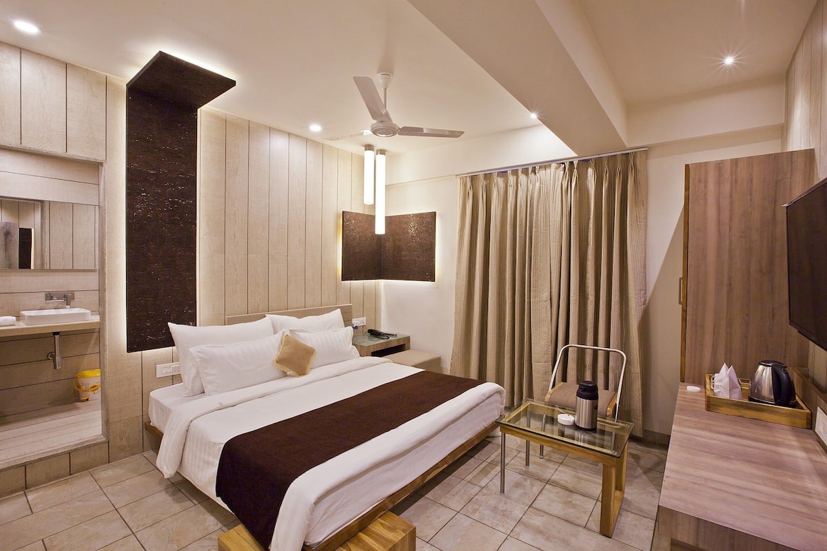 Quarto de hotel moderno com uma cama grande, mesa de cabeceira, escrivaninha, cadeira, televisão e banheiro com pia e espelho.