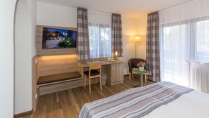 Quarto de hotel com cama, escrivaninha, cadeira, televisão fixada na parede e luminária.