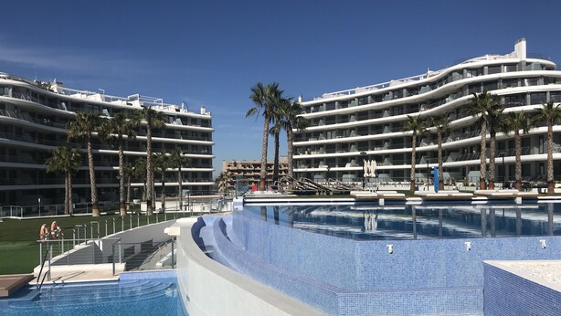 Um hotel moderno com uma grande piscina infinita, palmeiras e um céu azul límpido.