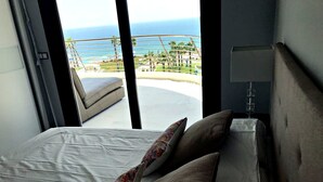 Uma cama com colchão branco e duas almofadas, uma mesa de cabeceira com abajur, e vista para o oceano e área de resort através de grandes janelas.