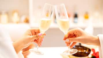 Duas mãos segurando taças de vinho, uma mesa branca e uma prateleira com várias garrafas ao fundo.