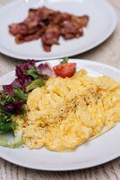 Um prato com ovos mexidos, acompanhado de folhas verdes e um tomate, e outro prato com bacon.