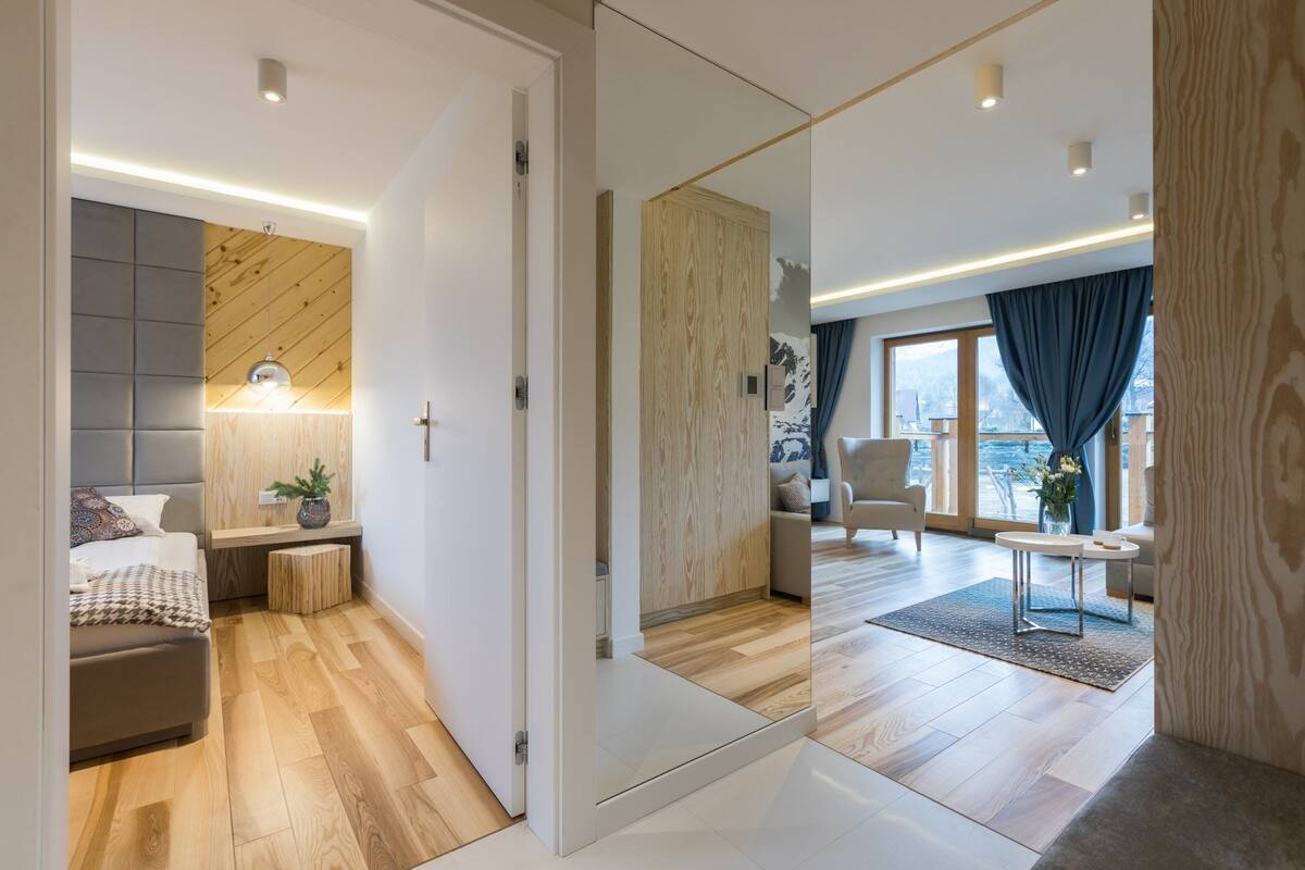 Quarto de hotel moderno com piso de madeira, cama com cabeceira, mesa de cabeceira com abajur e um espelho grande que reflete o ambiente.