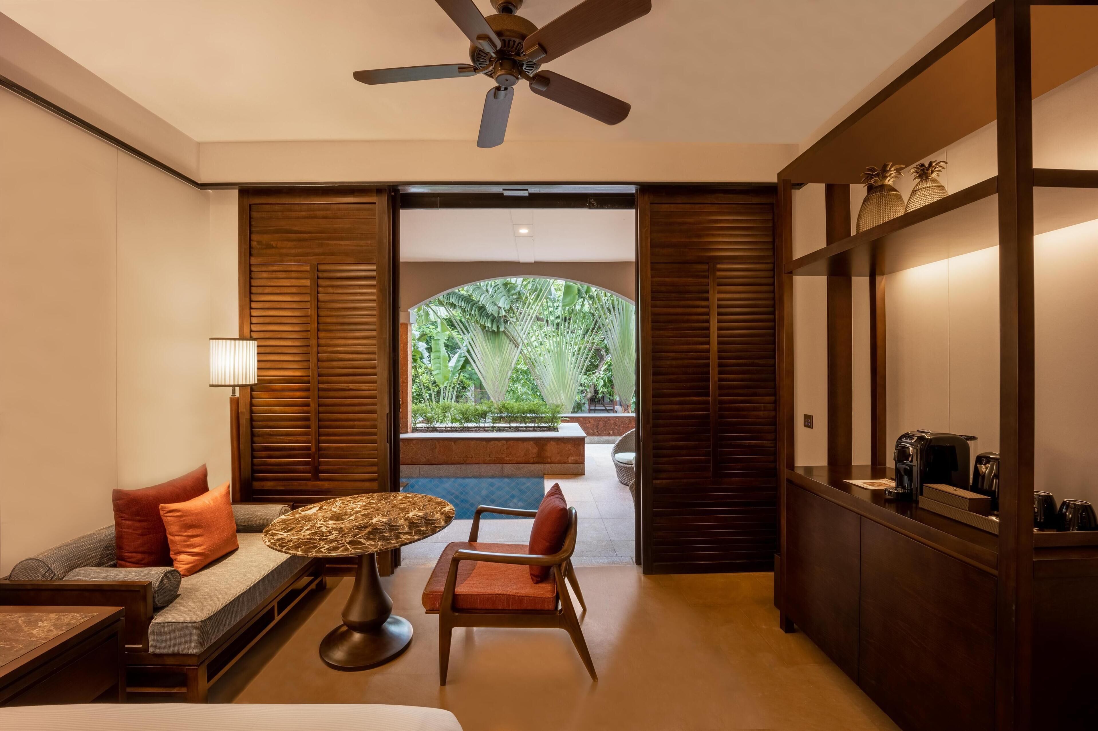 Quarto de hotel moderno com ventoinha no teto, sofá com almofadas, mesa redonda e vista para um jardim exterior através de uma porta de correr.
