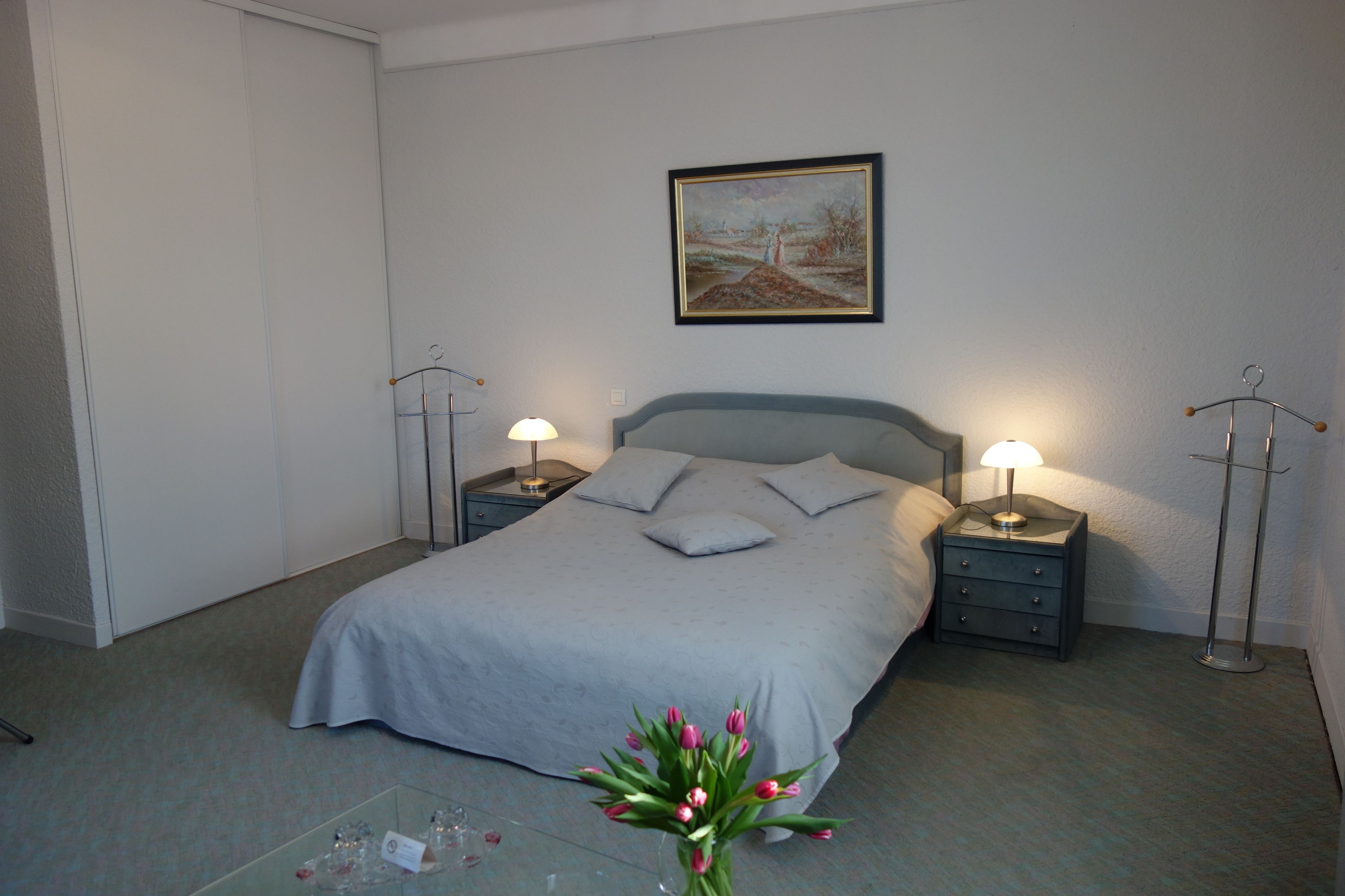 Quarto com cama, duas mesas de cabeceira com luminárias, um criado-mudo, um quadro na parede e um vaso com flores no chão.