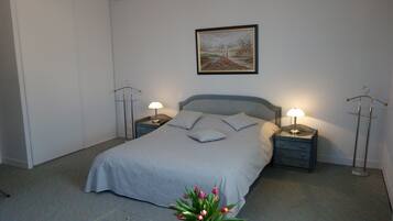 Quarto com cama, duas mesas de cabeceira com luminárias, um criado-mudo, um quadro na parede e um vaso com flores no chão.