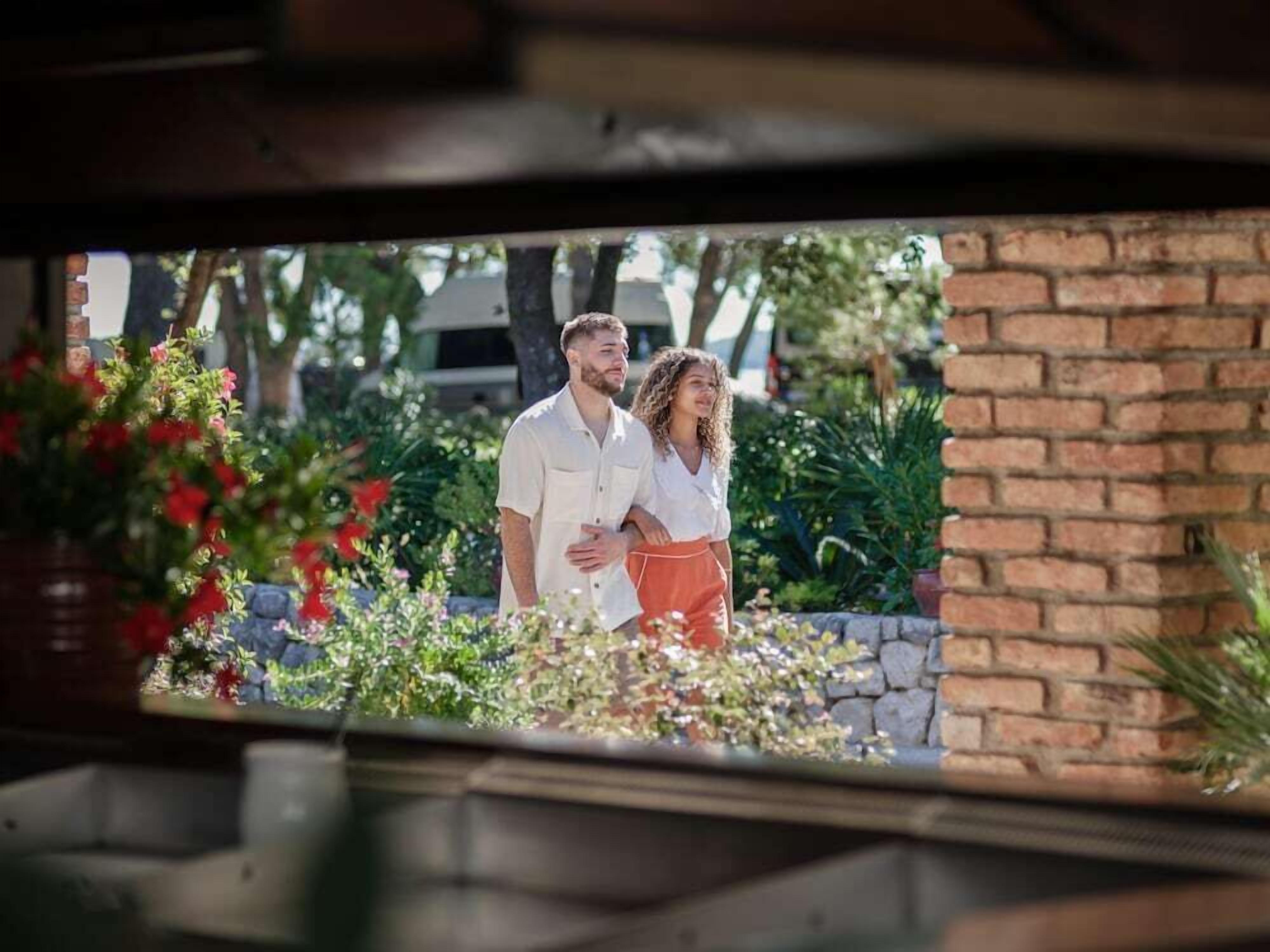 Um homem e uma mulher em um jardim, com um muro de tijolos e vegetação ao fundo.