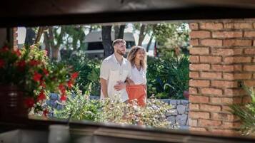 Um homem e uma mulher em um jardim, com um muro de tijolos e vegetação ao fundo.