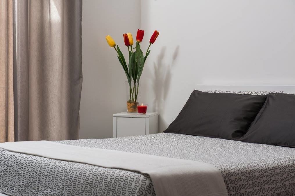 Uma cama bem arrumada com um cobre-leito estampado, uma mesa de cabeceira com um vaso de tulipas e uma mesinha de noite com uma vela.