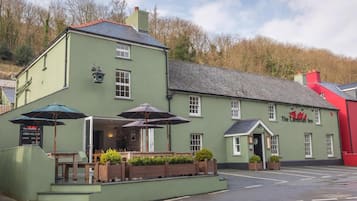 Um edifício verde com uma placa indicando 'The Cambrian Inn' e área externa com mesas e guarda-sóis.