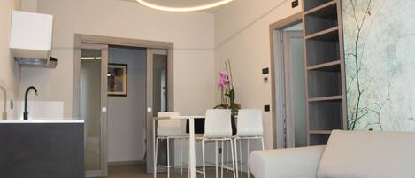 Uma sala de estar moderna com um sofá, uma mesa de centro de vidro, um espaço de refeições com cadeiras e uma cozinha com pia e armários.